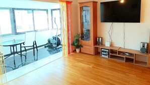 Uma sala de estar moderna com uma televisão grande, soalho de madeira, uma mesa de vidro e cadeiras, e uma vista através de janelas amplas.