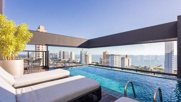 Piscina infinita no terraço com vista para a cidade, cadeiras de descanso e um vaso de planta.