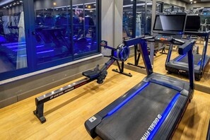 Ginásio com equipamentos de exercício, incluindo esteiras e um remo, com grandes janelas e piso de madeira.