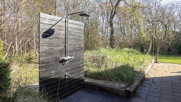 Estrutura de madeira com porta de metal e uma pequena janela, rodeada por vegetação natural e um caminho pavimentado.