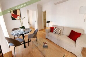 Uma sala de estar com sofá bege, almofadas vermelhas, uma mesa de centro de vidro e um vaso com planta.