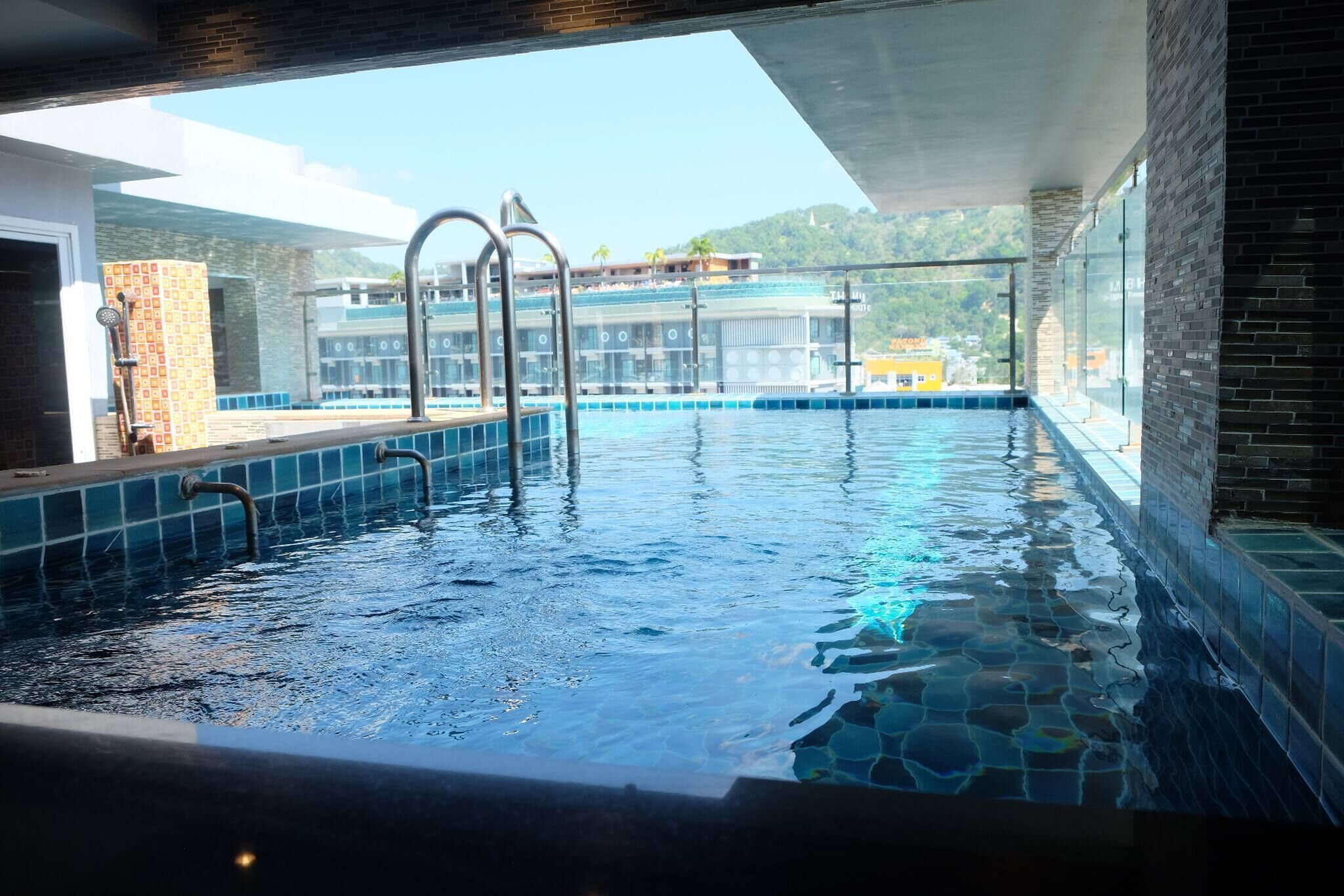 Piscina interior com água azul cristalina, escadas de metal e vista para um edifício e colinas ao fundo.