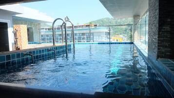 Piscina interior com água azul cristalina, escadas de metal e vista para um edifício e colinas ao fundo.