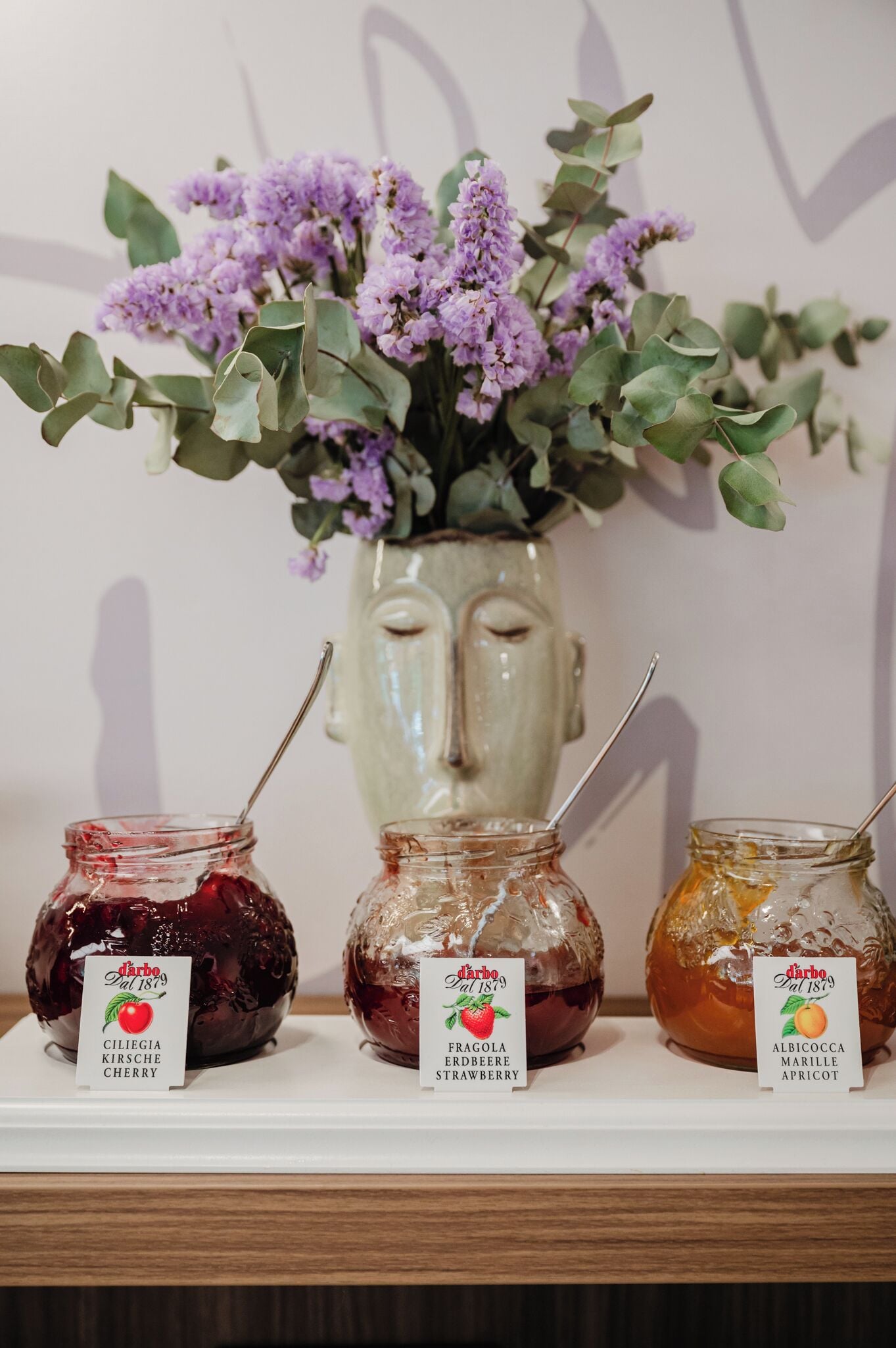 Três potes de geleia com etiquetas, um vaso com flores roxas e uma escultura facial de cerâmica.
