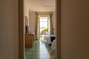 Quarto de hotel com cama, escrivaninha e cadeira. Há uma janela com cortinas e vista para o verde lá fora.