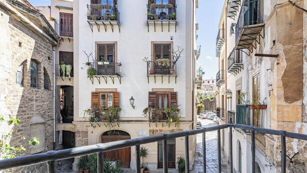 Uma rua estreita com edifícios de ambos os lados, com varandas adornadas com plantas em vasos, paredes de pedra e um caminho de paralelepípedos.