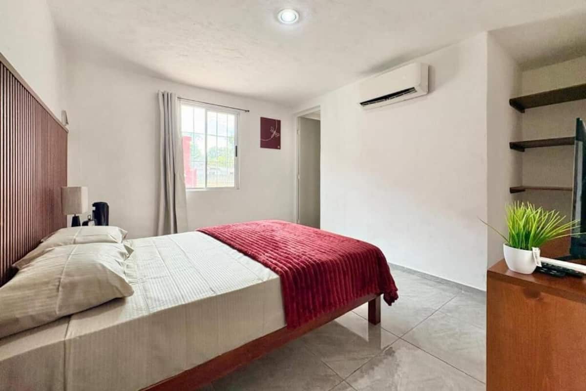 Quarto de hotel com cama, escrivaninha, planta em vaso e aparelho de ar condicionado.