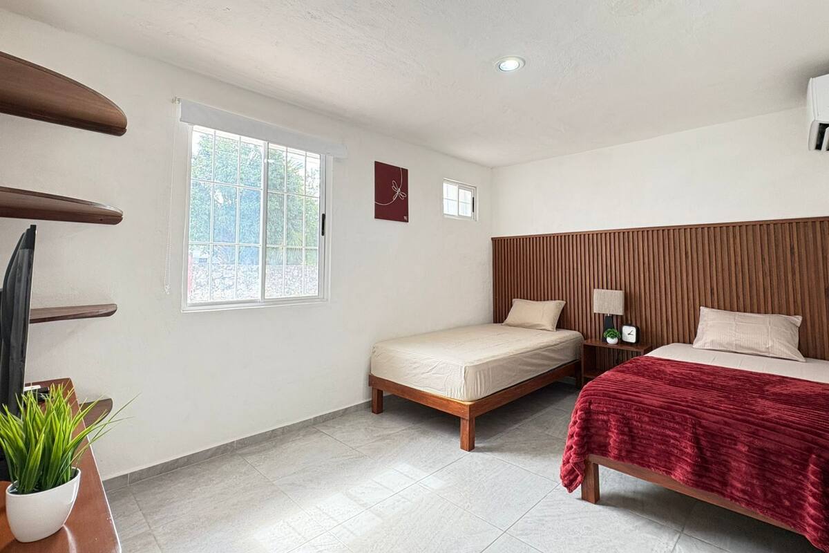 Quarto com duas camas, uma janela, um vaso de planta e um relógio na parede.