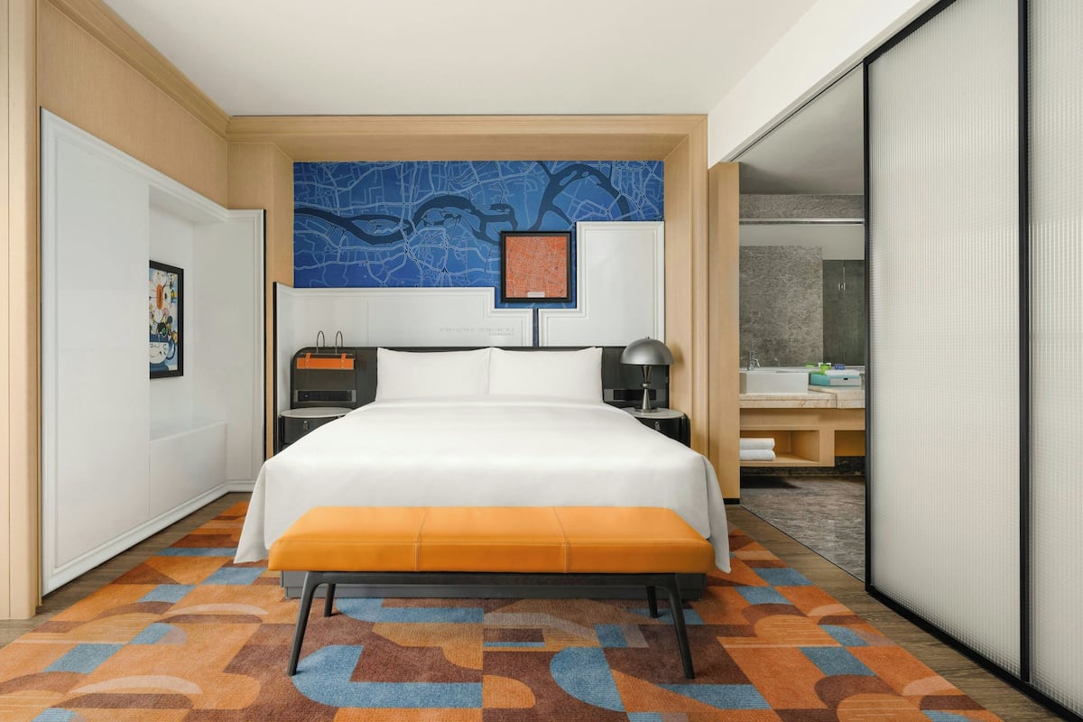 Quarto de hotel com cama, mesa de cabeceira, luminária, banheiro com pia e um quadro abstrato grande na parede.