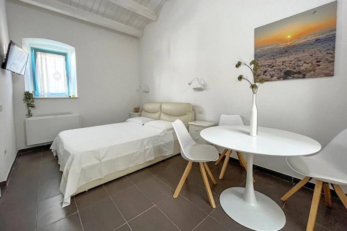 Quarto com cama, mesa de cabeceira, cadeira, pequena mesa, janela com cortinas, luminária na parede, pintura de um pôr do sol e planta em vaso.
