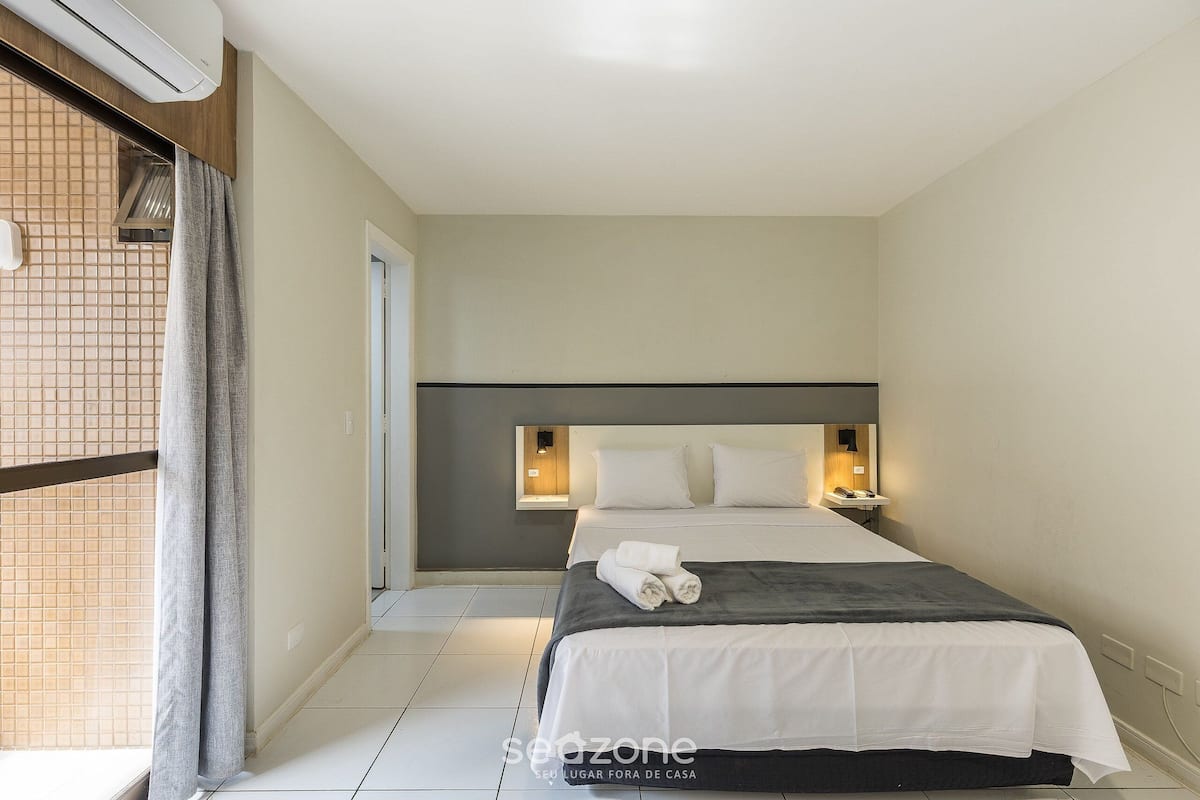 Quarto de hotel com cama, mesa de cabeceira, luminária na parede e janela com cortinas.
