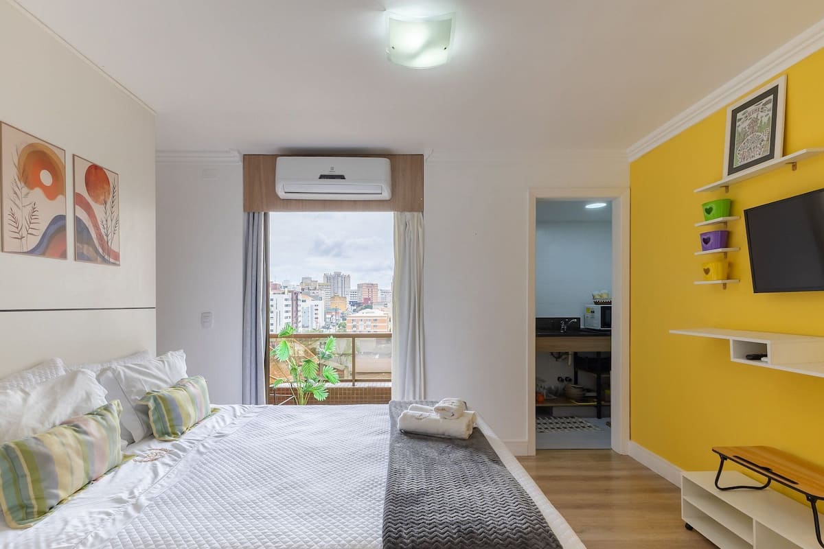 Quarto com cama, janela com vista para a cidade, TV e área de cozinha.