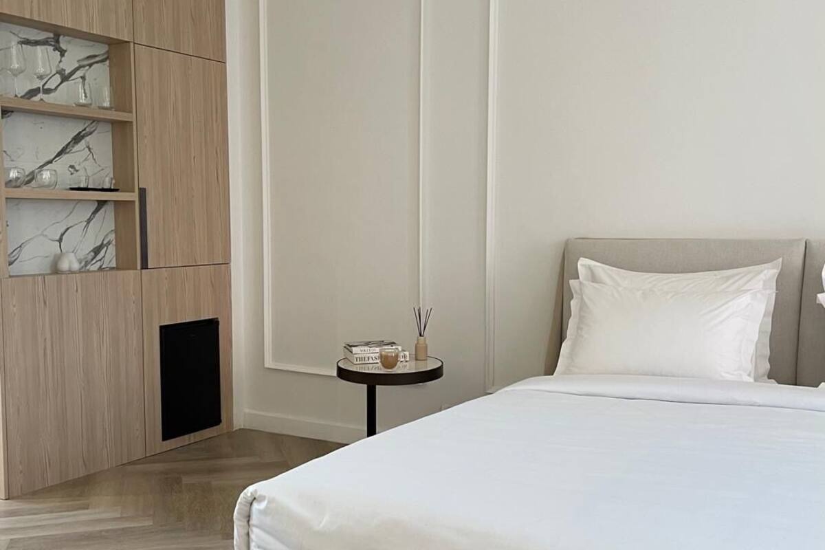 Quarto de hotel moderno com uma cama grande, uma mesinha auxiliar, uma prateleira embutida de madeira com copos e uma parede branca.