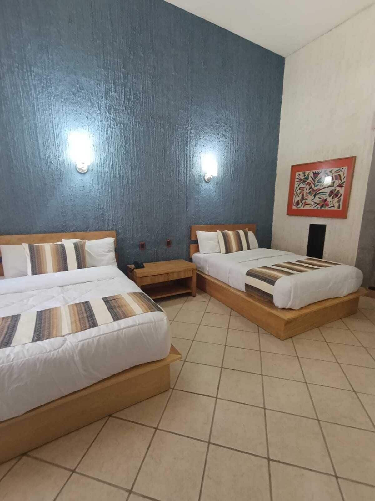 Quarto de hotel com duas camas, uma mesinha de cabeceira de madeira, uma luminária fixa na parede e um quadro emoldurado.