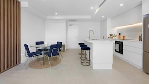 Uma cozinha moderna com armários brancos, uma ilha central com banquetas e uma área de refeições com cadeiras azuis e uma mesa de vidro.