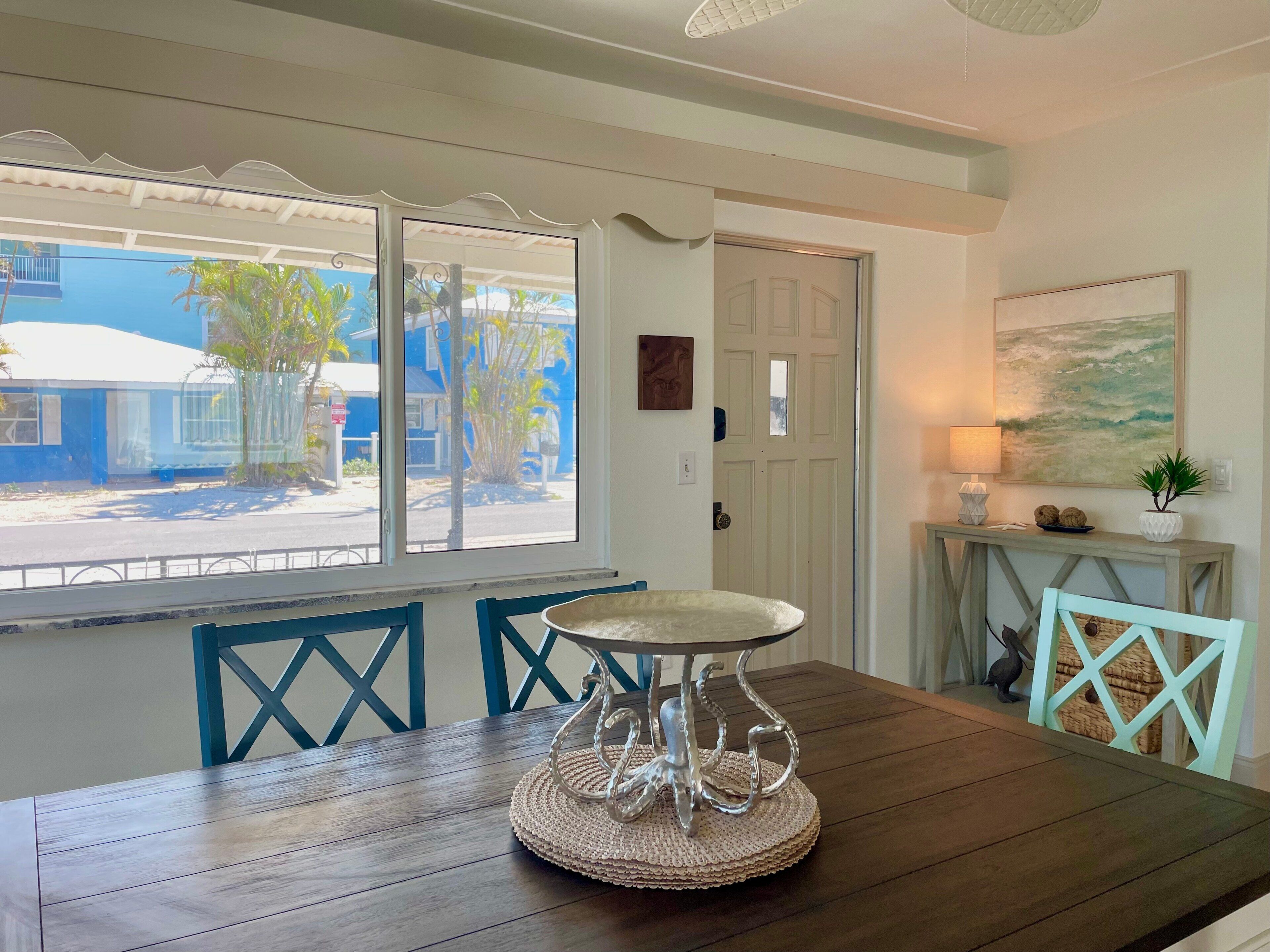 Uma área de refeições com mesa de madeira, duas cadeiras azuis, base de mesa metálica decorativa, um jogo americano trançado, uma porta com moldura branca, um quadro pendurado na parede, um pequeno console com uma luminária e vista para um prédio azul e palmeiras lá fora.