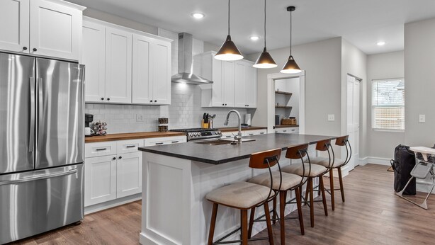 Cozinha moderna com armários brancos, eletrodomésticos de aço inoxidável, ilha central com banquetas, iluminação pendente e vista para a área de jantar adjacente.