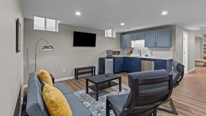 Uma sala de estar moderna com sofá, poltrona e mesa de centro. Cozinha com armários azuis e micro-ondas visível ao fundo.