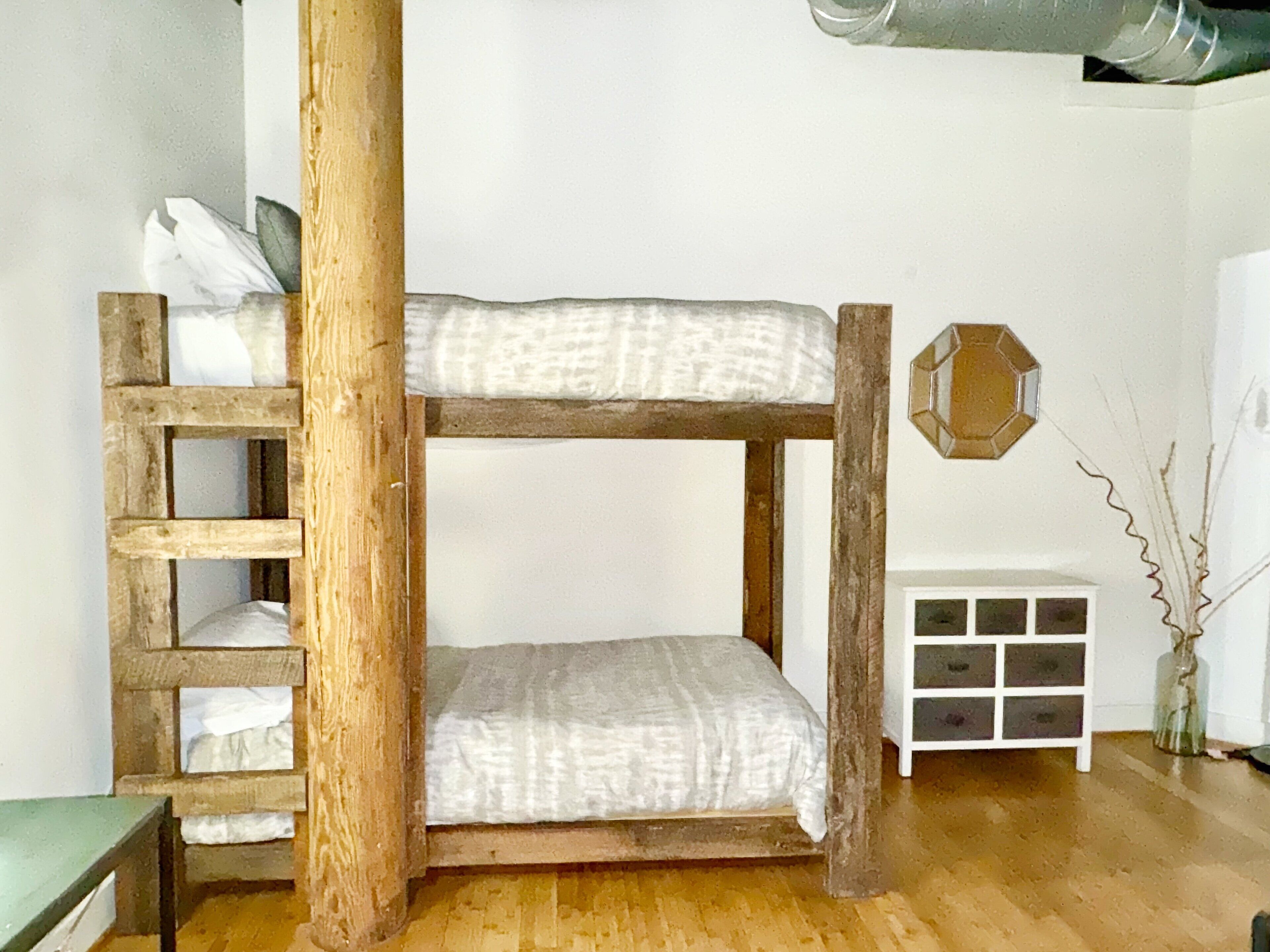 Uma cama beliche com escada de madeira, um móvel de armazenamento branco e um espelho hexagonal na parede.