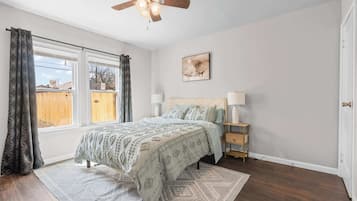 Quarto com cama, mesas de cabeceira, ventilador de teto e janela com cortinas.