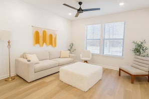 Uma sala de estar moderna com sofá, pufes e poltronas, um ventilador de teto e uma janela grande com persianas.