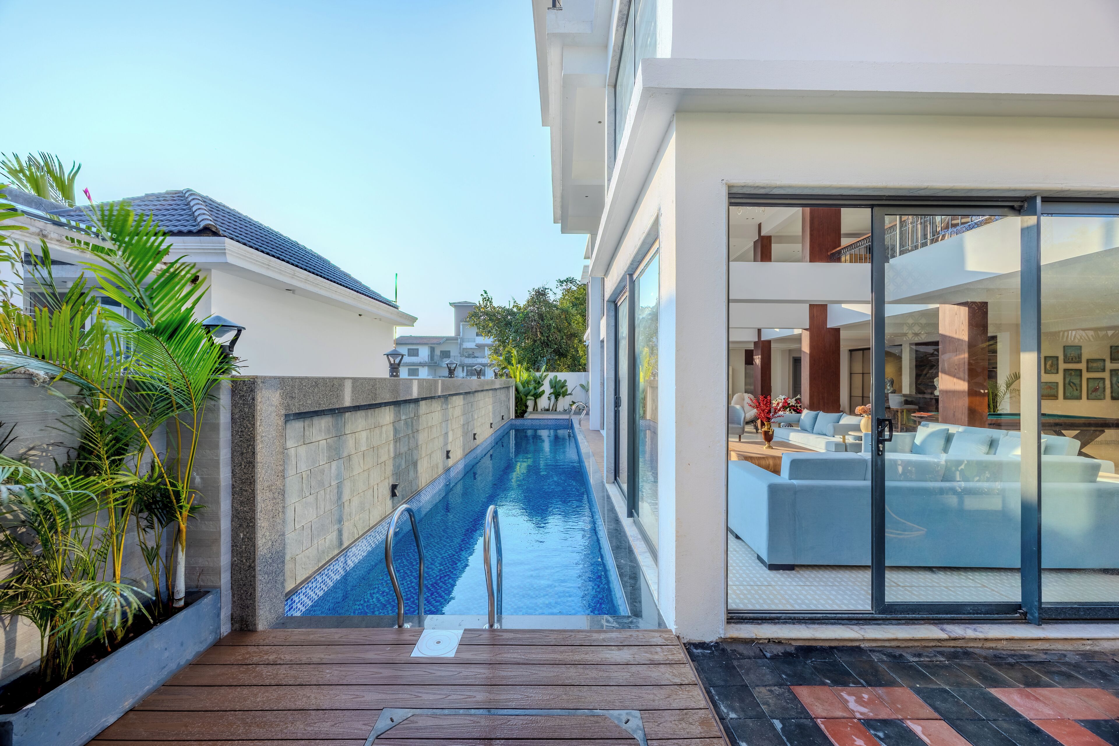 Uma área de piscina moderna com um deck de madeira, uma piscina azul cristalina e um espaço de estar envidraçado com um sofá e uma mesa de centro.