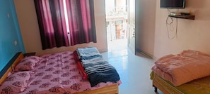 Um quarto com cama, um banco, uma televisão na parede e uma janela com cortinas.