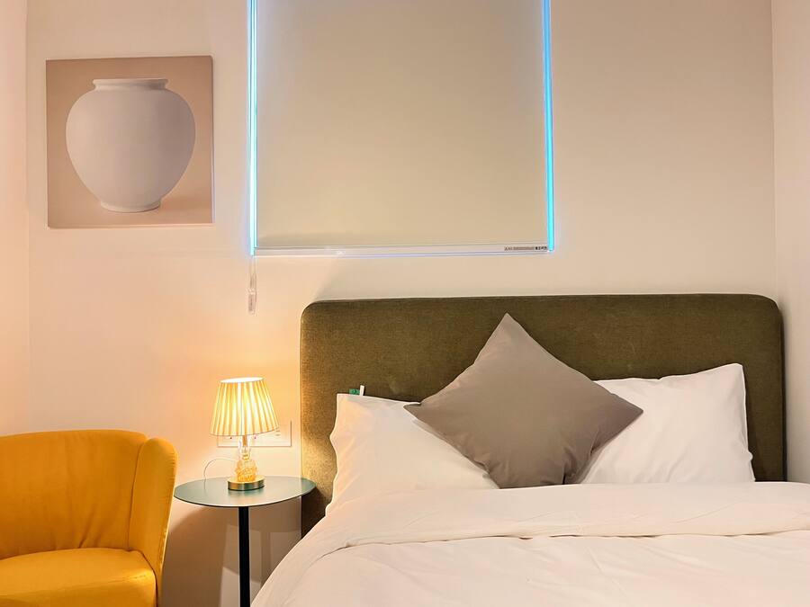 Quarto de hotel com uma cama, mesa de cabeceira com abajur, cadeira e um vaso fixado na parede.