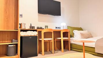 Quarto de hotel com uma cama, uma escrivaninha com duas cadeiras, um pequeno frigorífico, uma TV fixada na parede e um cesto de lixo.
