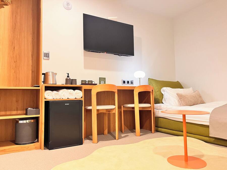 Quarto de hotel com uma cama, uma escrivaninha com duas cadeiras, um pequeno frigorífico, uma TV fixada na parede e um cesto de lixo.