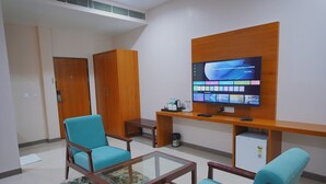 Uma sala de estar moderna com televisão de tela plana na parede, uma escrivaninha de madeira, duas poltronas azuis, uma mesa de centro de vidro e um frigorífico pequeno.