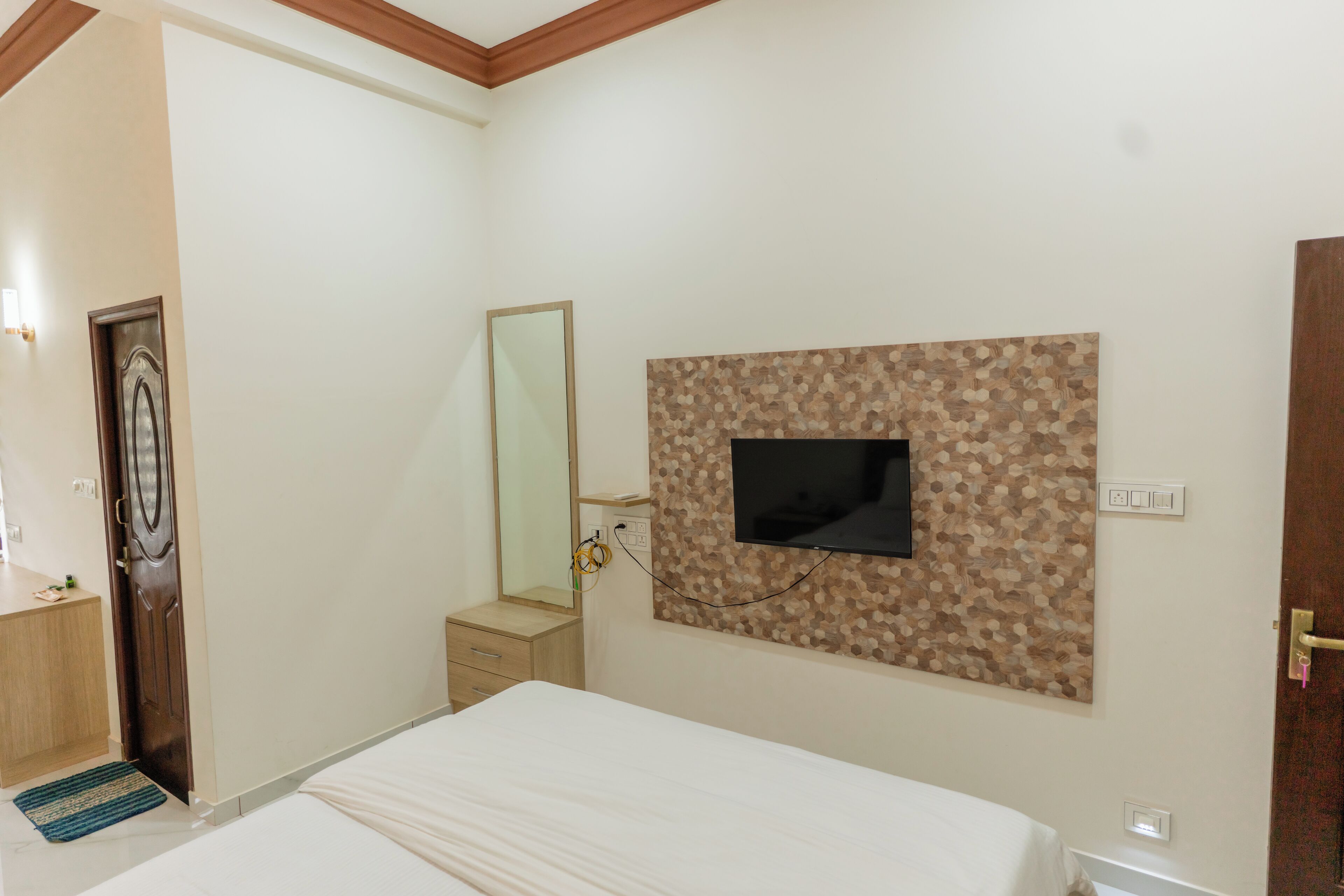 Quarto com cama, porta de madeira, cômoda, espelho e televisão fixada em parede texturizada.