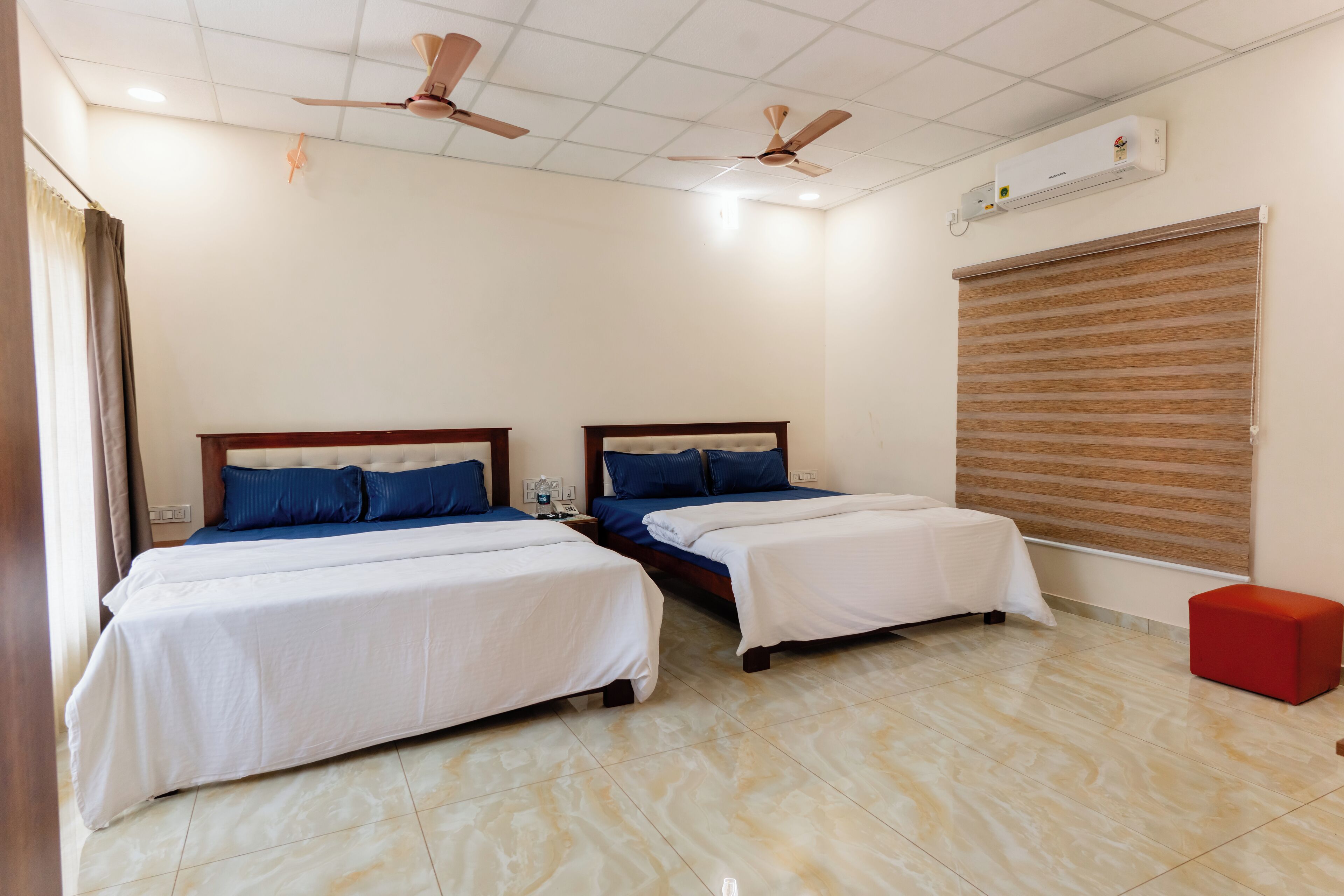 Quarto de hotel com duas camas, ventiladores de teto, ar condicionado, um banquinho vermelho e uma janela grande com cortinas.