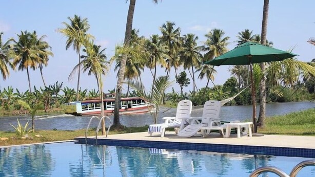 Piscina com espreguiçadeiras e guarda-sol, cercada por palmeiras e um barco ao fundo.