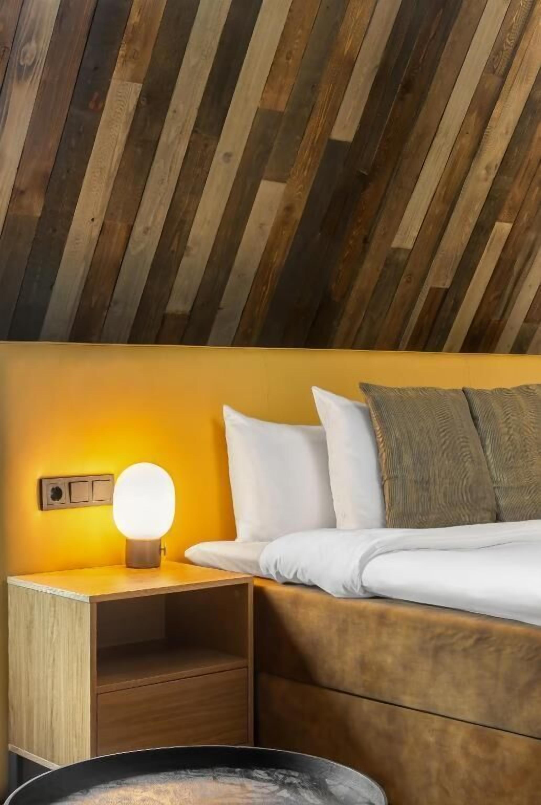 Quarto com teto de ripas de madeira, mesa de cabeceira com abajur, cama com roupa de cama branca e uma pequena mesa redonda.