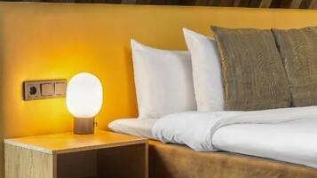 Quarto com teto de ripas de madeira, mesa de cabeceira com abajur, cama com roupa de cama branca e uma pequena mesa redonda.