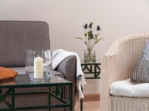 Um quarto com uma mesa redonda de metal verde, uma vela branca, um vaso de vidro com flores, uma cadeira de vime e um sofá cinza.