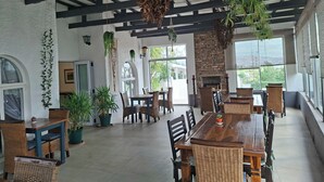 Uma sala de jantar ampla, com uma longa mesa de madeira e cadeiras de vime, cercada por plantas em vasos e folhagem pendente.