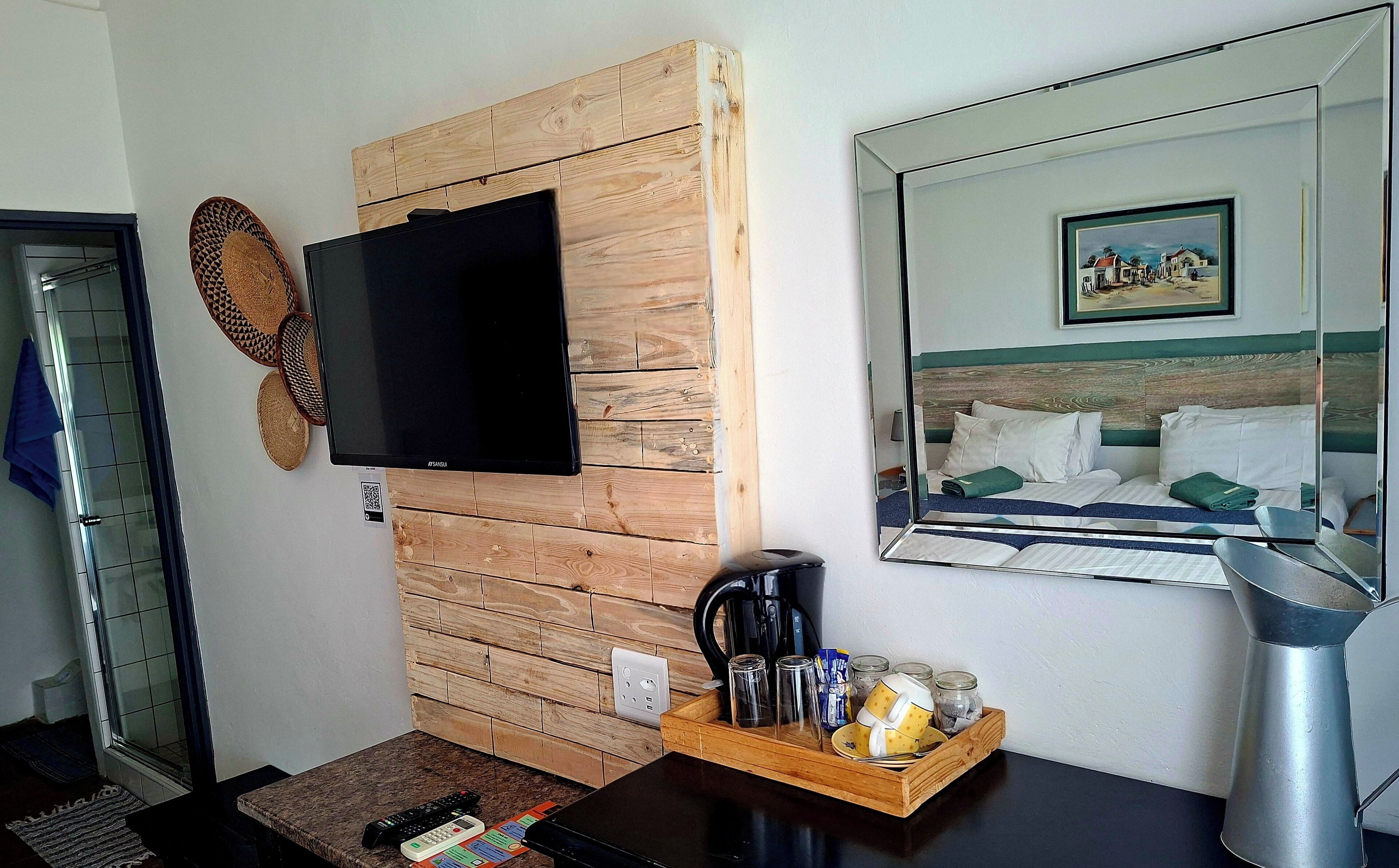 Quarto de hotel com painel de parede de madeira, televisão de tela plana, cama com cabeceira verde, mesa de cabeceira com cafeteira, copos e potes.