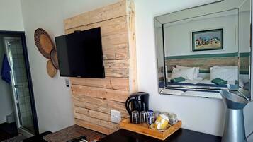 Quarto de hotel com painel de parede de madeira, televisão de tela plana, cama com cabeceira verde, mesa de cabeceira com cafeteira, copos e potes.