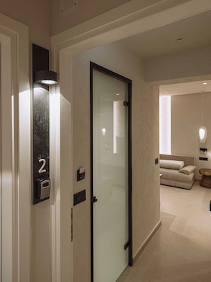 Um corredor moderno com porta de vidro, placa com o número '2', leitor de cartão-chave e área de estar minimalista com sofá e mesa redonda de madeira.