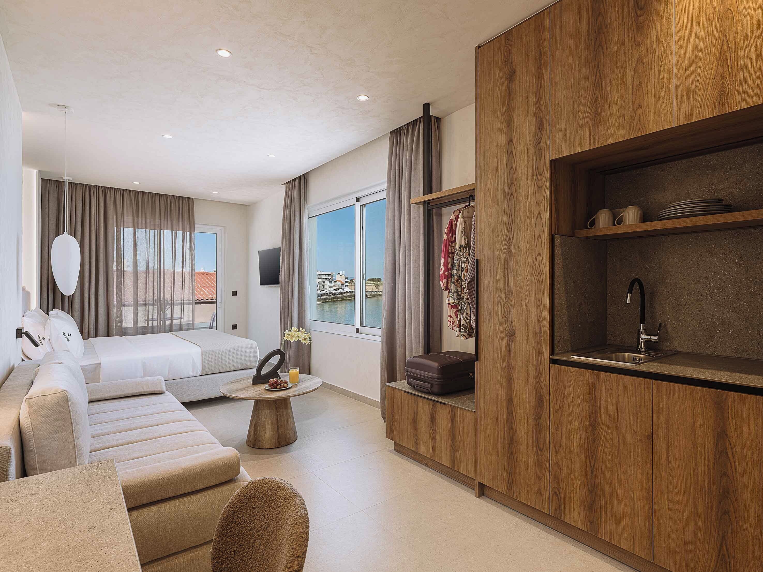 Quarto de hotel moderno com cama, sofá, área de refeições e vista para o mar.