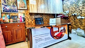 Balcão de recepção de hotel com bancada de madeira, uma imagem emoldurada de divindades e um aviso com detalhes sobre comodidades como estacionamento gratuito e acesso à internet.
