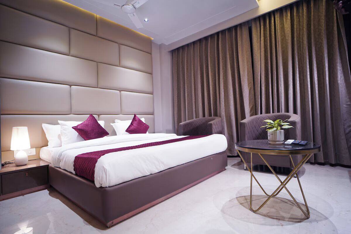 Quarto de hotel moderno com uma cama grande, uma mesa de cabeceira com luminária, uma mesinha redonda com uma planta e uma janela grande com cortinas.