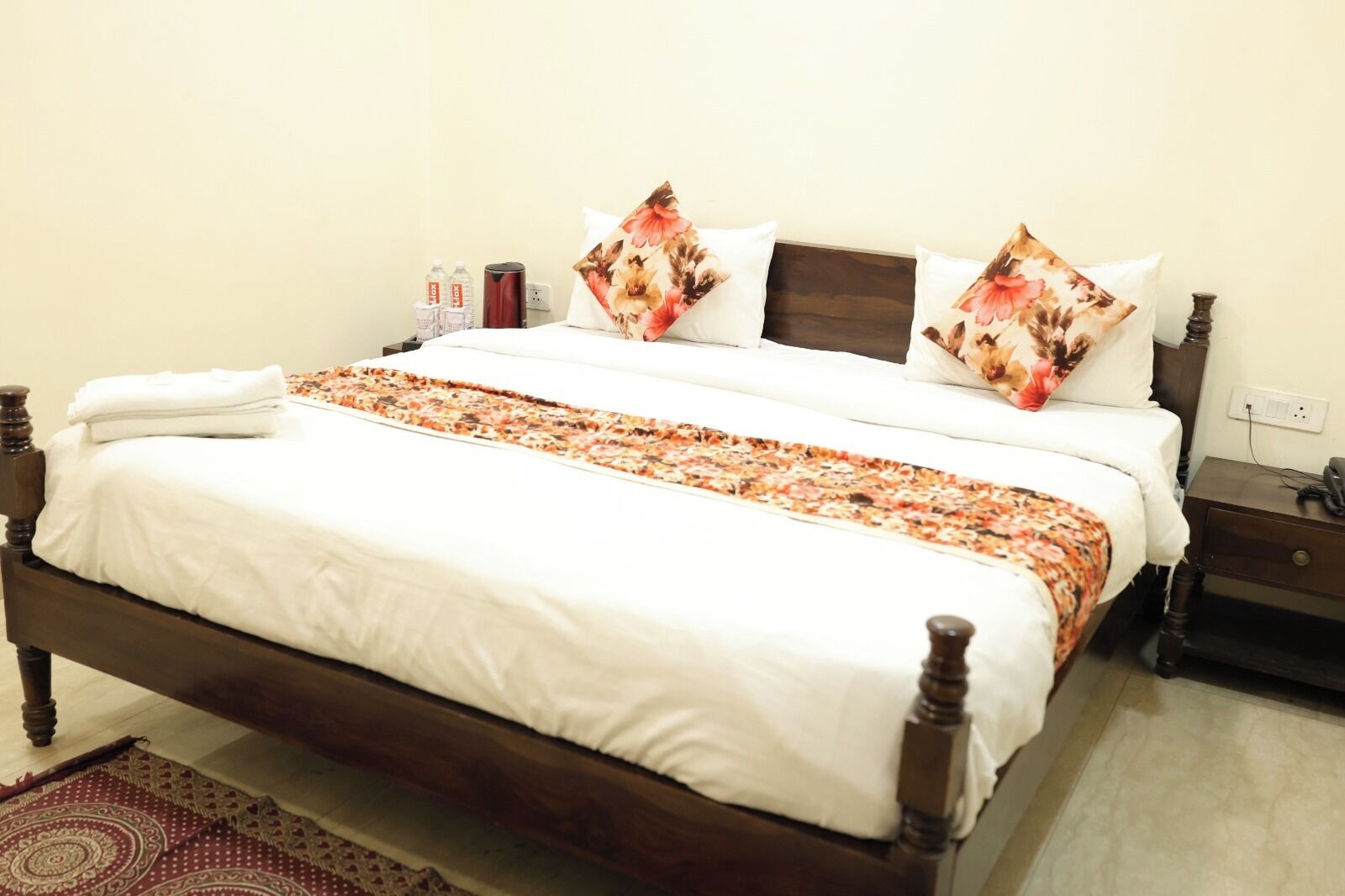 Uma cama bem arrumada com lençóis brancos e um manta com padrão floral, uma mesa de cabeceira de madeira com abajur, e uma pequena mesa com um telefone.