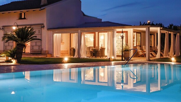 Uma área à beira da piscina com um espaço coberto para sentar, piscina e uma casa com fachada branca e telhado escuro.
