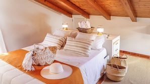 Cama bem arrumada com lençóis brancos e almofadas listradas, um cesto de vime no criado-mudo e um chapéu branco sobre a cama.