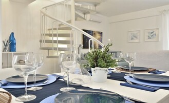 Mesa de jantar posta com pratos azuis, copos de vinho transparentes e um vaso branco com folhagem verde.