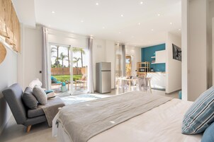 Quarto de hotel moderno com uma cama grande, área de estar com sofá e poltrona, cozinha compacta e porta de vidro deslizante que dá para uma varanda com mobiliário de exterior.