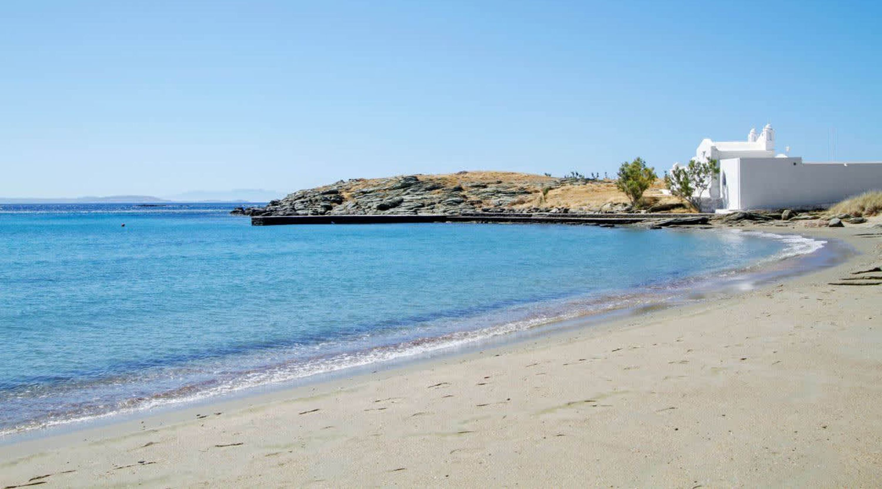 Uma praia com águas azul-claras, areal arenoso e um edifício branco com um afloramento rochoso nas proximidades.